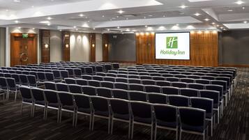 Uma sala de conferências com fileiras de cadeiras, uma tela de projeção exibindo o logotipo do Holiday Inn e uma parede de madeira com uma placa de saída.