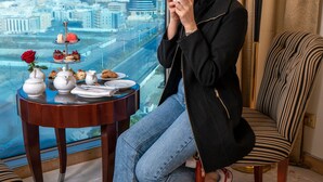 Mulher sentada perto de uma janela com vista para a cidade, uma mesa com um conjunto de chá e um abajur.