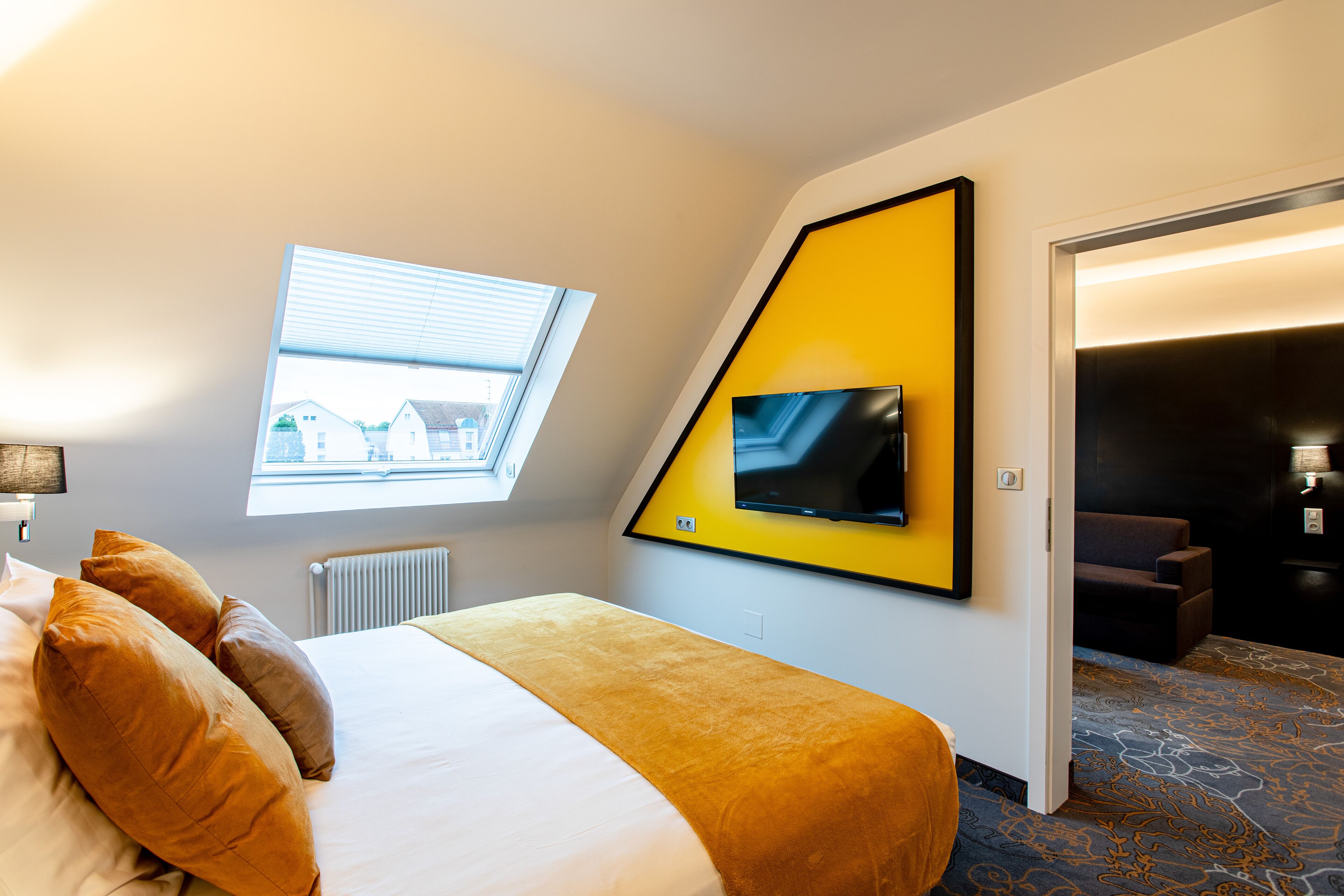 Quarto de hotel com uma cama, televisão fixada na parede, janela e porta que dá para outro espaço.