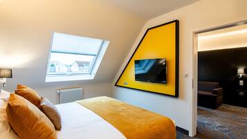 Quarto de hotel com uma cama, televisão fixada na parede, janela e porta que dá para outro espaço.