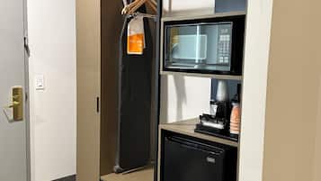 Quarto compacto com área de cozinha compacta, incluindo micro-ondas, máquina de café e frigorífico. Possui um pequeno espaço de estar com um banco e uma sacola pendurada.