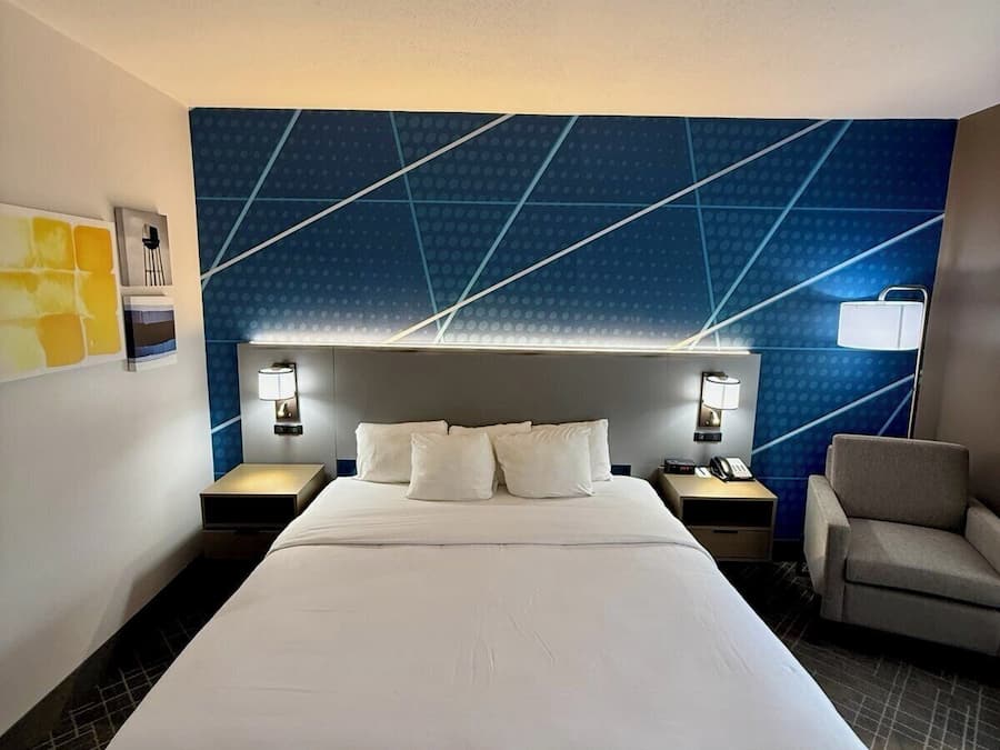 Quarto de hotel com uma cama grande, duas mesas de cabeceira com abajures, uma cadeira e parede com padrão geométrico.