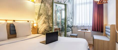 Quarto de hotel moderno com uma cama grande, um laptop sobre a cama, uma mesa pequena, uma cadeira e um espelho.