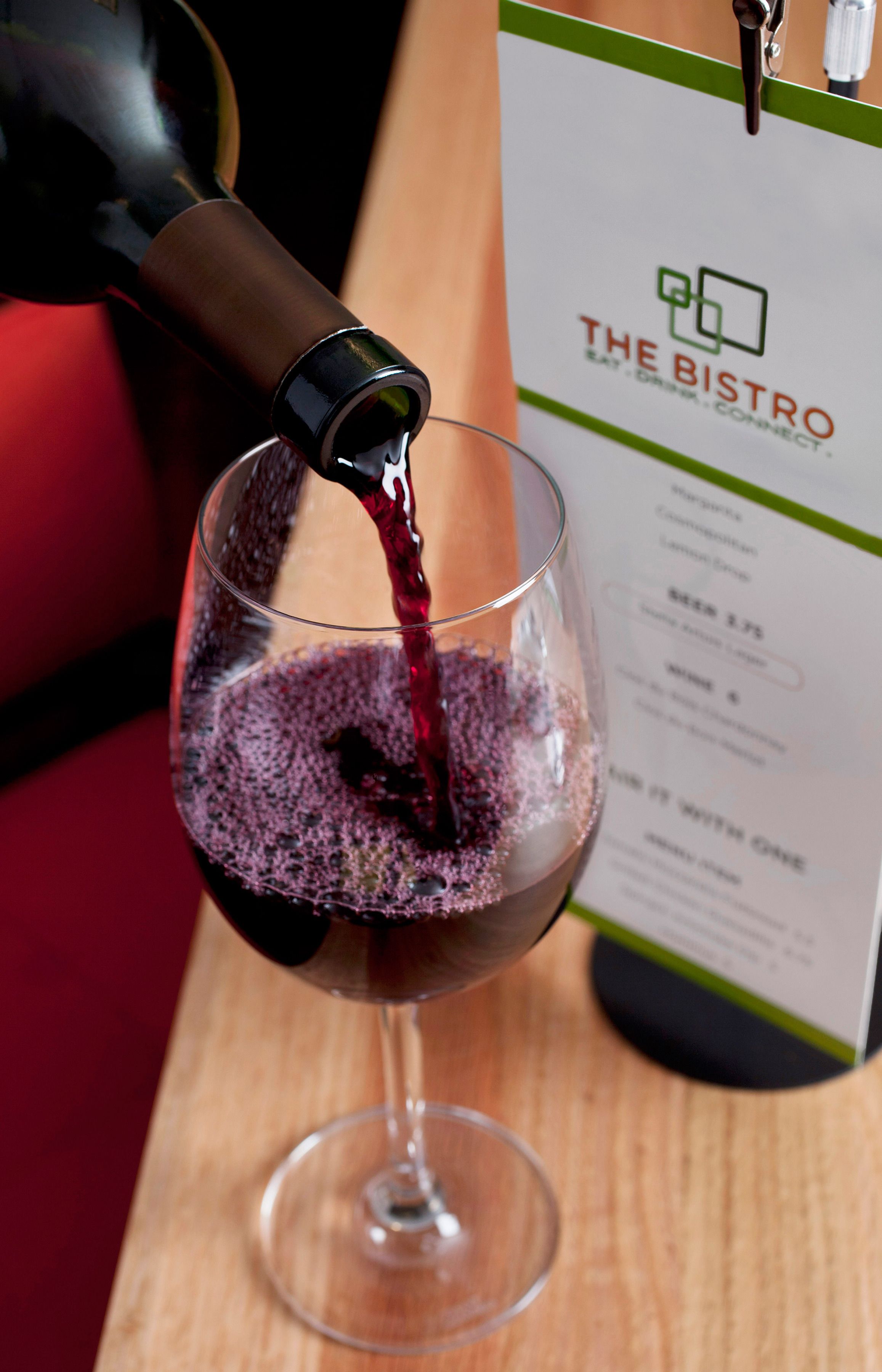 Uma taça de vinho sendo servida com vinho tinto, um cardápio e uma garrafa de vinho sobre uma mesa de madeira.