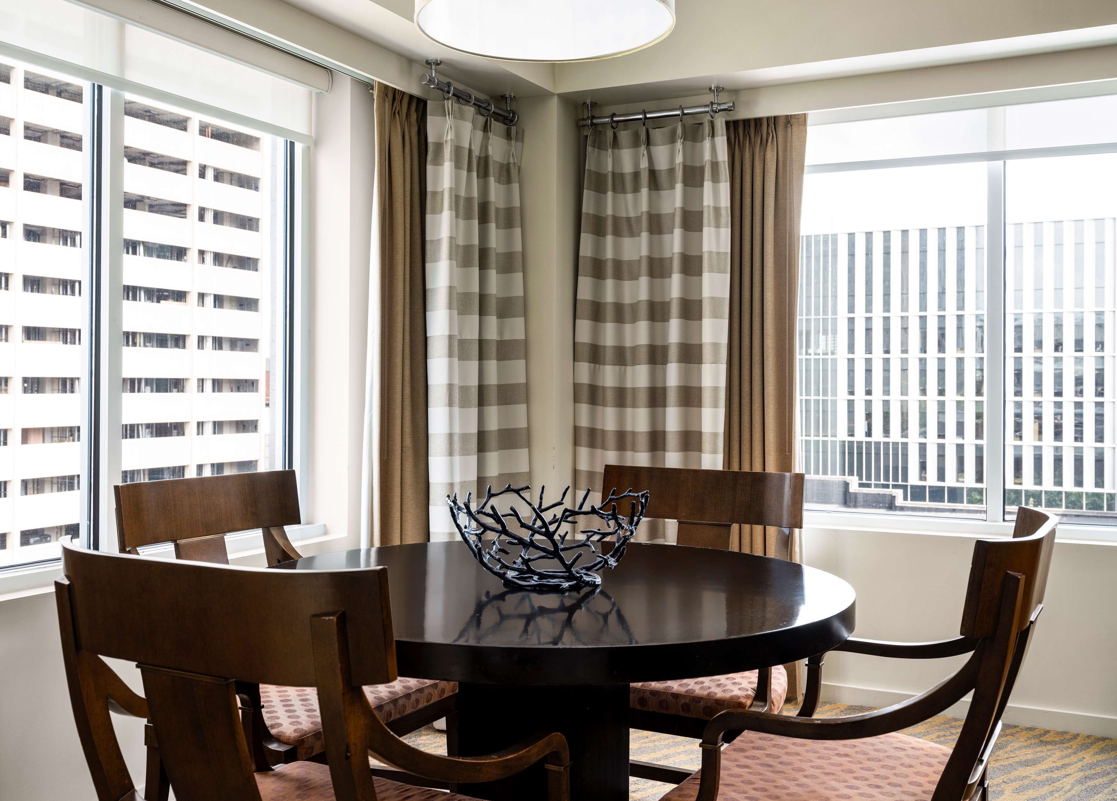 Uma área de refeições no canto, com uma mesa redonda, quatro cadeiras de madeira, uma tigela de vidro decorativa e cortinas.