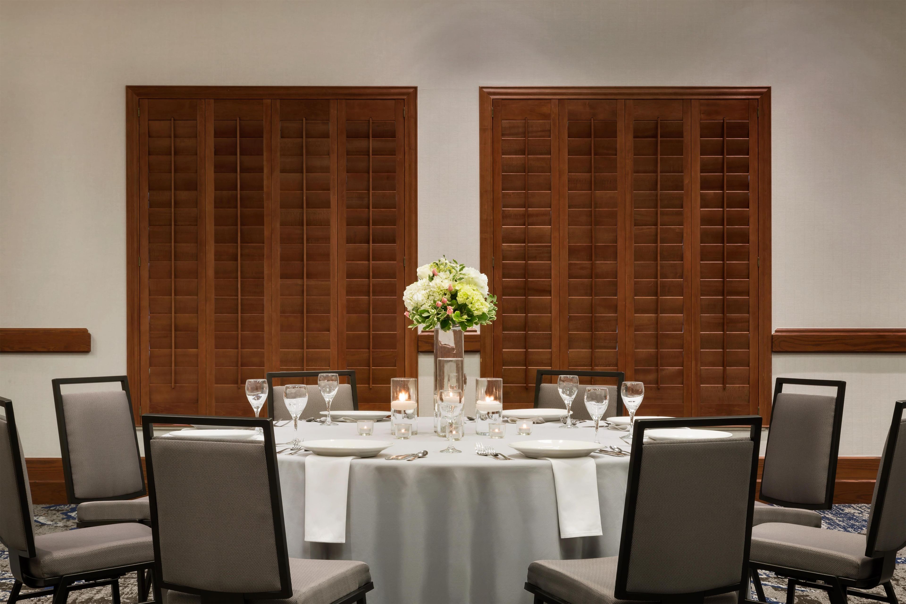 Sala de conferência com mesa redonda para seis pessoas, limitada por duas paredes revestidas de madeira, e seis cadeiras com estofamento cinza.
