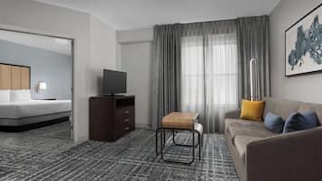 Quarto de hotel moderno com carpete cinza estampado, um sofá com almofadas, um banquinho de madeira, uma TV em um móvel e uma grande janela com cortinas cinzas.