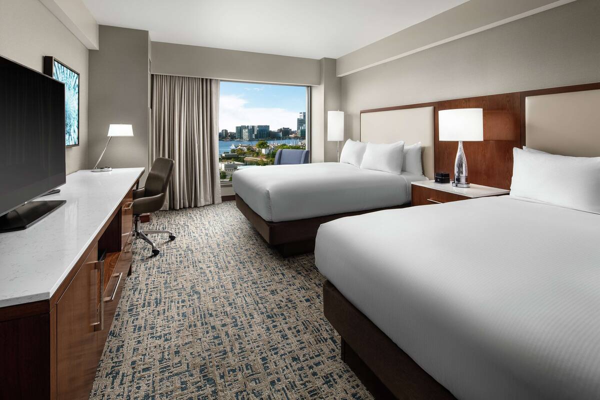 Quarto de hotel com uma cama grande, uma cama menor, uma mesa com televisão e vista para a cidade através de uma janela.
