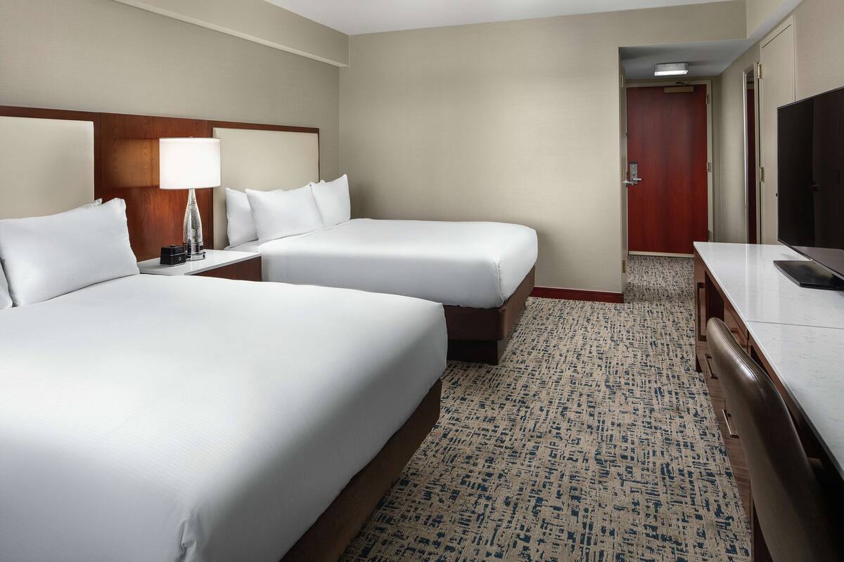 Quarto de hotel com duas camas, uma escrivaninha com televisão de ecrã plano, uma lâmpada e um tapete estampado.
