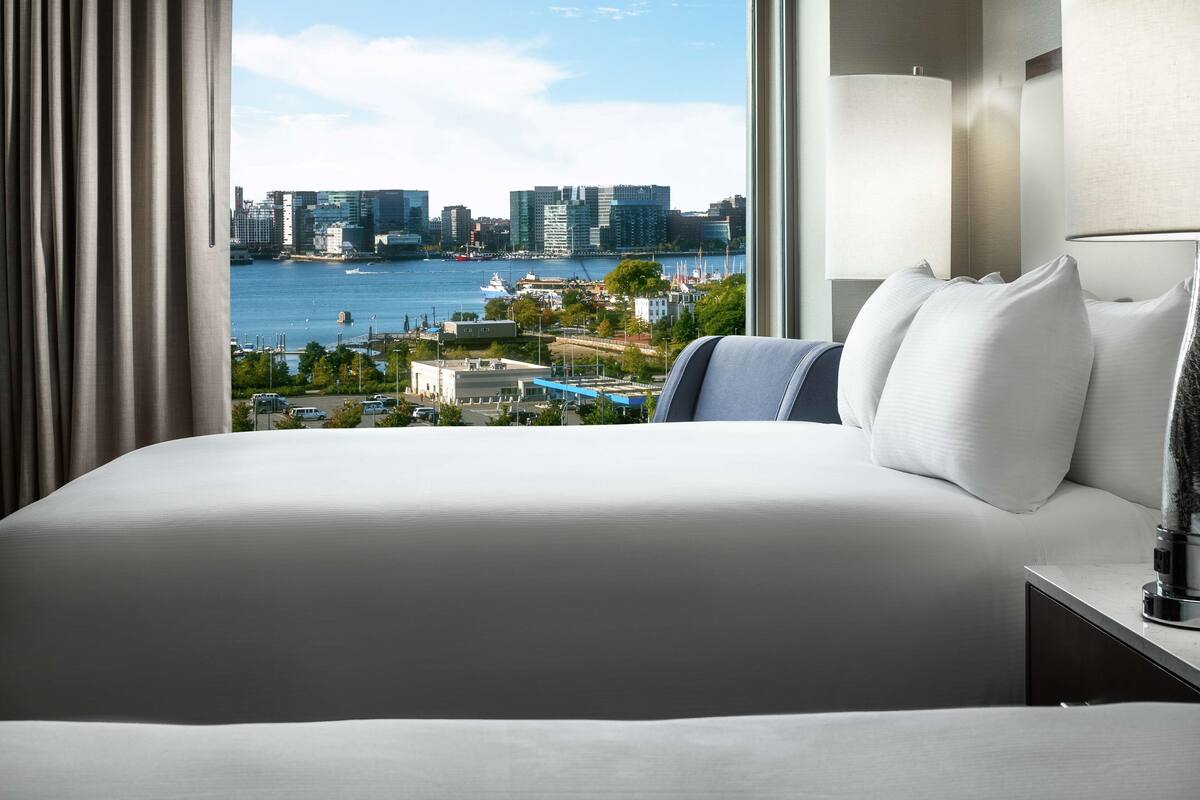 Quarto de hotel com uma cama grande, vista para a paisagem urbana e para a água, e um abajur na mesa de cabeceira.