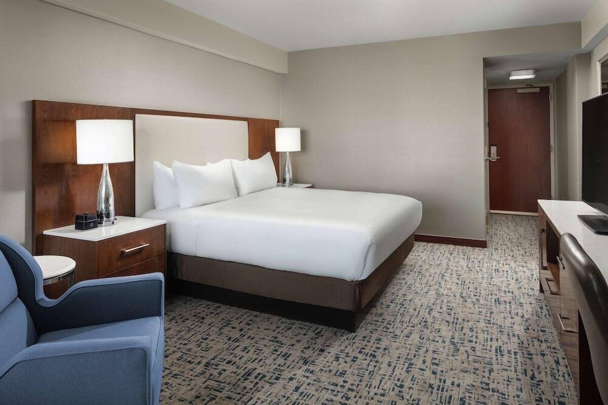 Quarto de hotel com uma cama grande, duas mesas de cabeceira com luminárias, uma escrivaninha com cadeira e uma televisão.