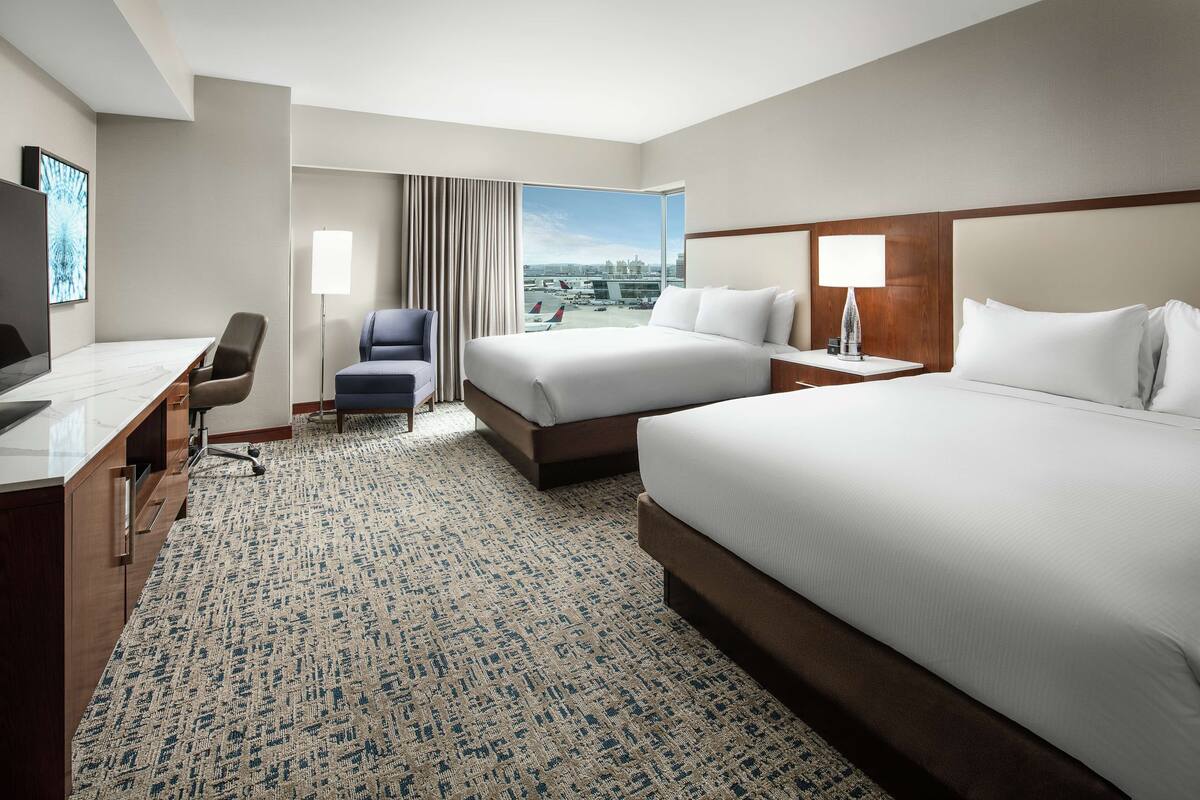 Quarto de hotel com duas camas, uma escrivaninha, uma cadeira, uma TV e uma janela grande com vista para a cidade.