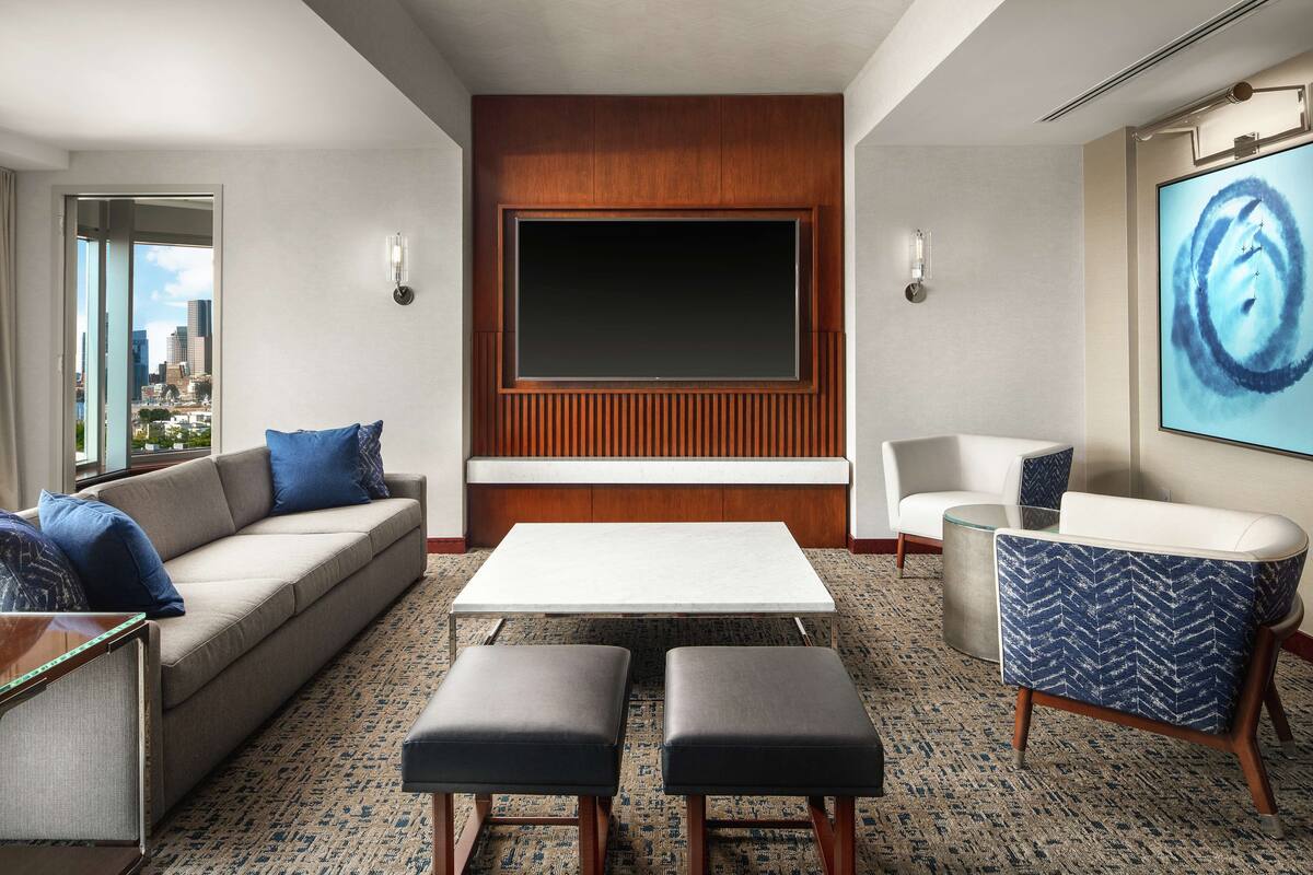 Quarto de hotel moderno com uma TV de tela plana grande presa a um painel de madeira, um sofá com almofadas azuis, uma mesa de centro branca, dois pufes pretos, um tapete estampado e vista para a cidade por uma janela grande.