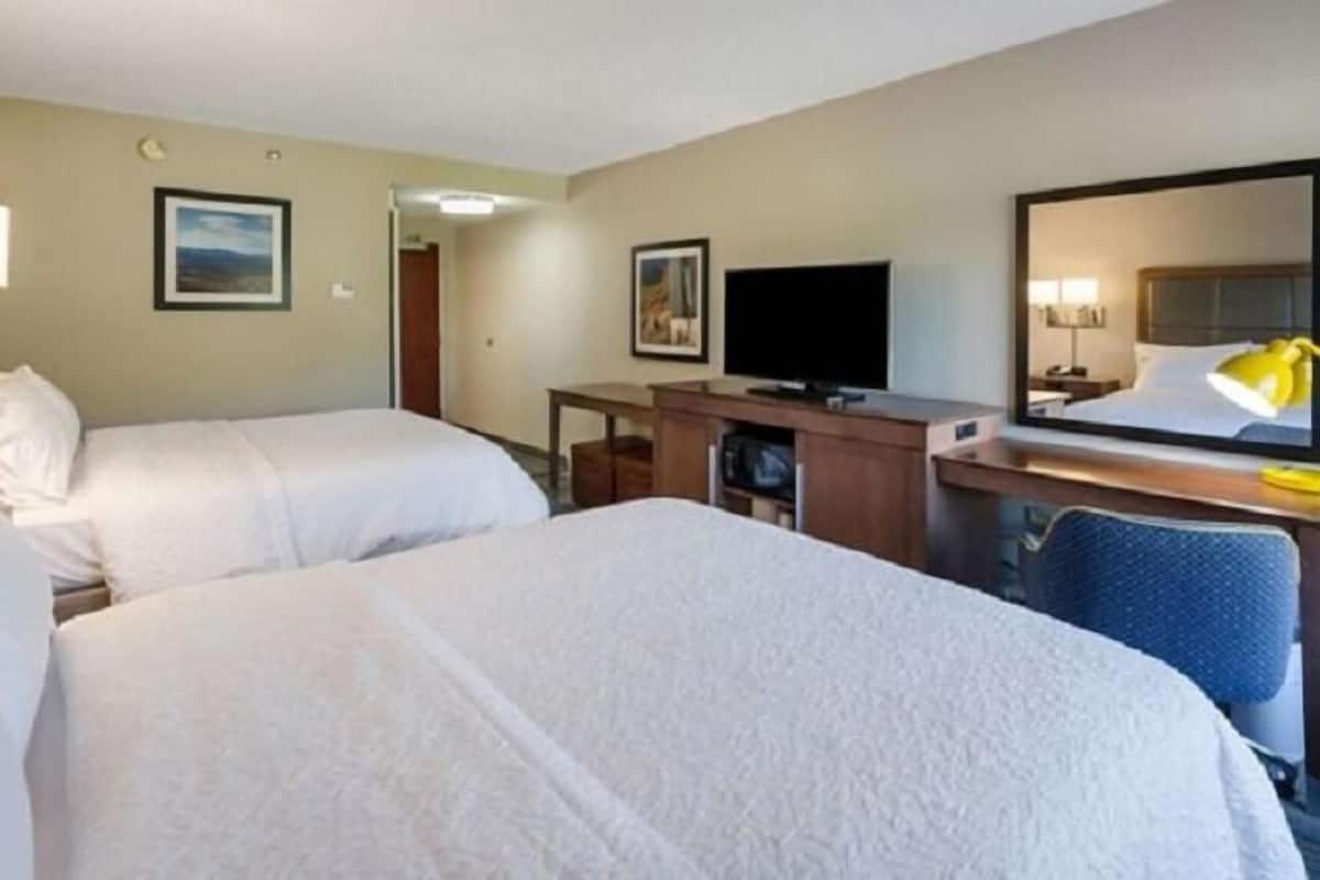 Quarto de hotel com duas camas, uma escrivaninha, televisão de tela plana, espelho e luminária.