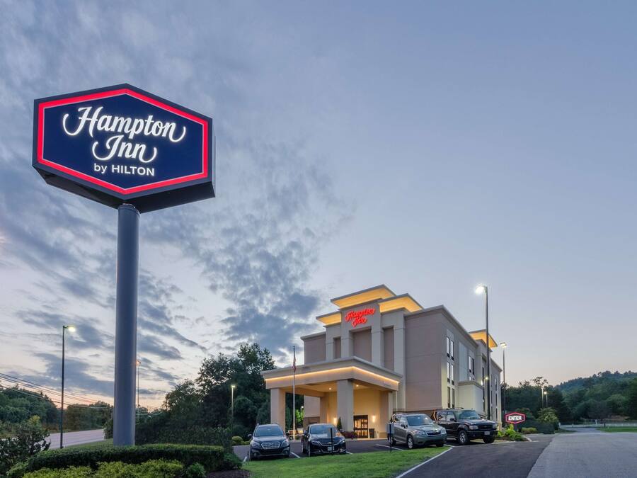 Um hotel Hampton Inn by Hilton com uma placa grande, um estacionamento com vários carros e um gramado bem cuidado.