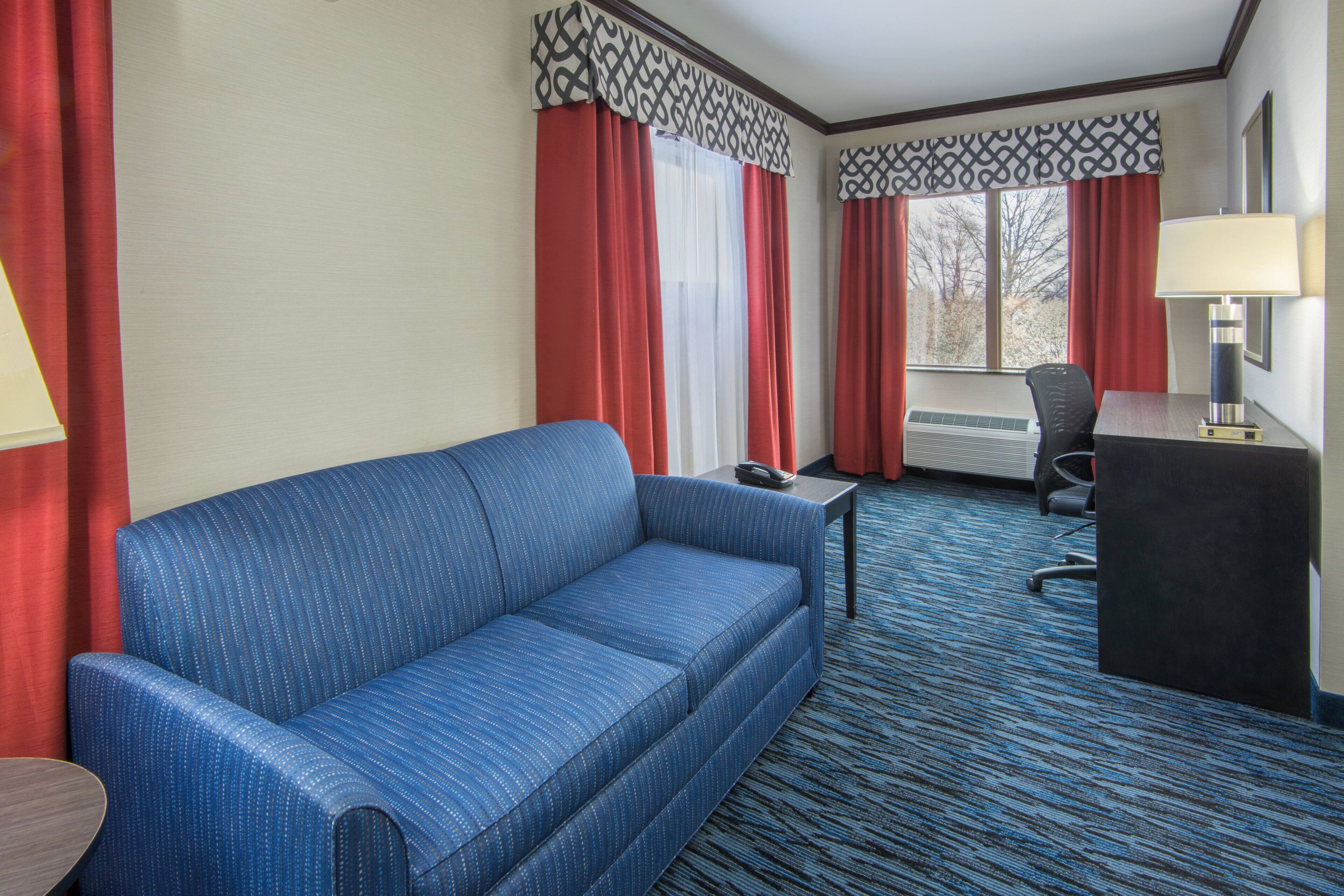 Quarto de hotel com sofá azul, escrivaninha com luminária e vista para árvores pela janela.