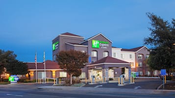 Edifício do Holiday Inn Express & Suites com design moderno, exibindo um logotipo verde e branco, um estacionamento e um mastro com a bandeira americana.