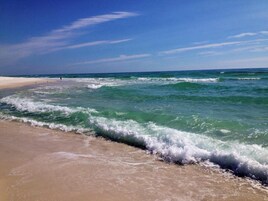 Uma praia com ondas avançando em direção à costa, céu claro e uma pessoa distante caminhando na areia.