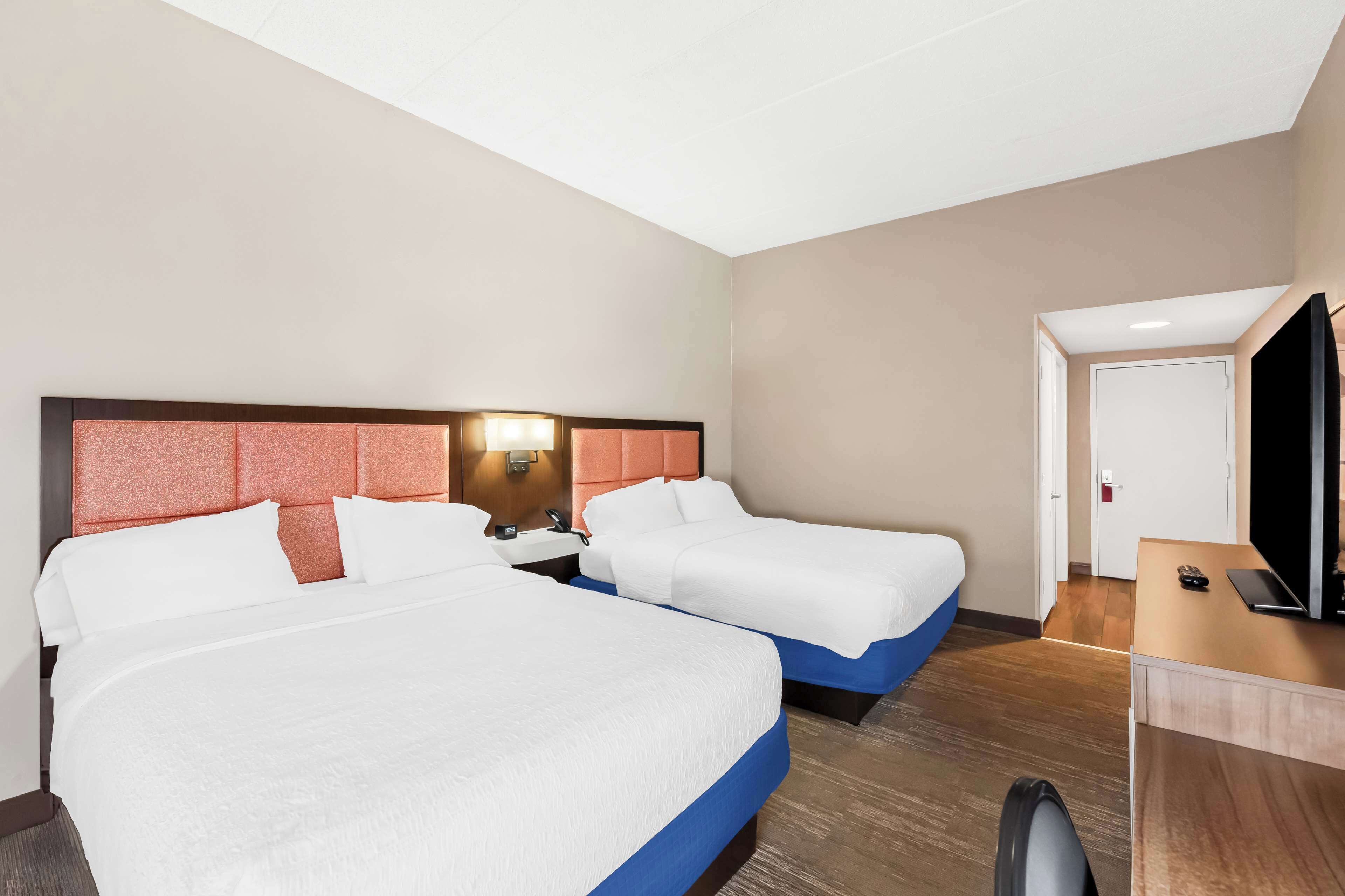 Quarto de hotel com duas camas, uma mesa, uma televisão e uma porta.