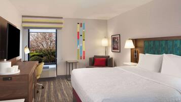 Quarto de hotel com cama, escrivaninha, cadeira, televisão e vista para uma árvore pela janela.