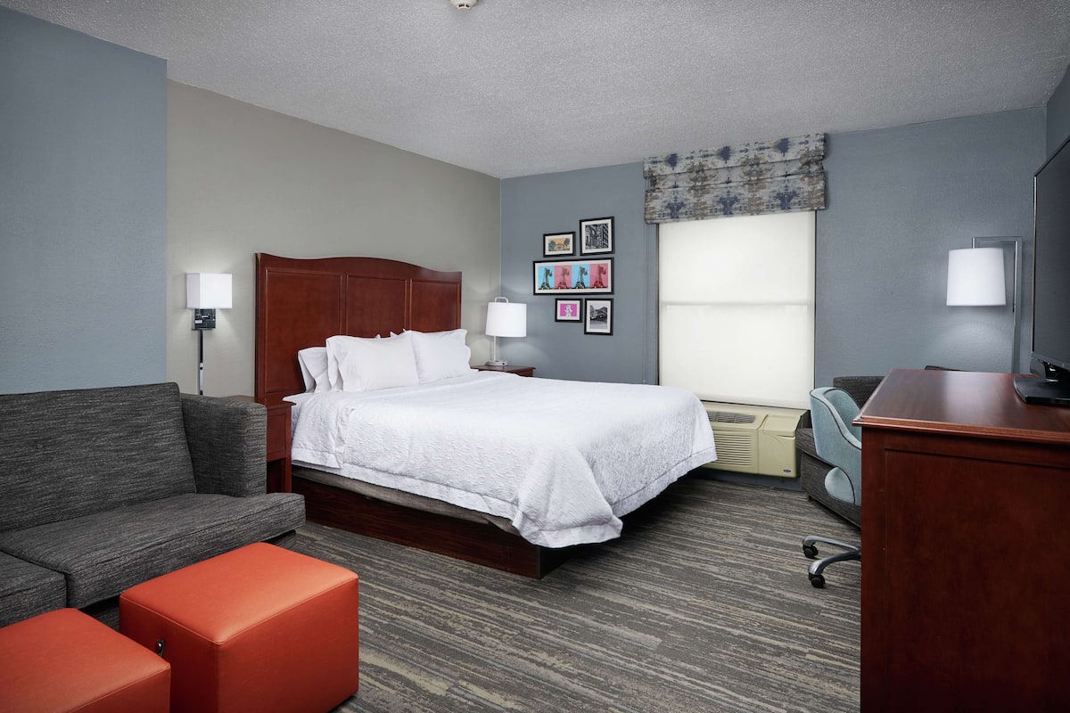 Quarto de hotel com cama, sofá, puf, escrivaninha e cadeira.