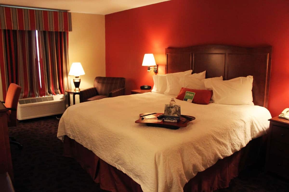Quarto de hotel com uma cama grande, duas luminárias de cabeceira, uma cadeira, uma mesa e uma janela com cortinas.