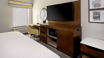 Quarto de hotel com cama, escrivaninha, televisão de tela plana, micro-ondas, cadeira e janela grande com persianas.