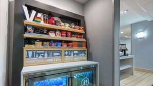 Máquina de vendas com lanches e bebidas, uma pequena geladeira com água engarrafada e uma prateleira com diversos produtos embalados.