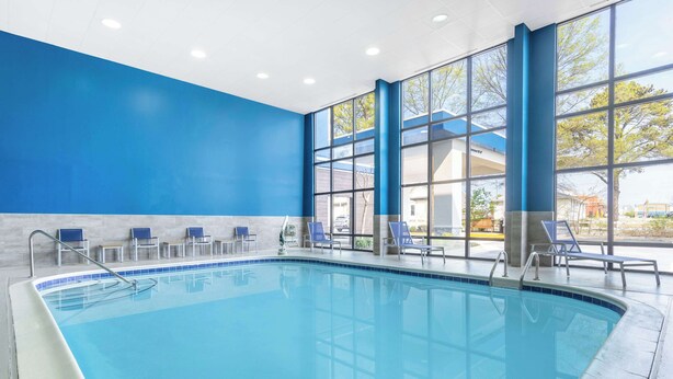 Piscina interna com água azul límpida, cercada por cadeiras azuis e grandes janelas que oferecem vista para o exterior.