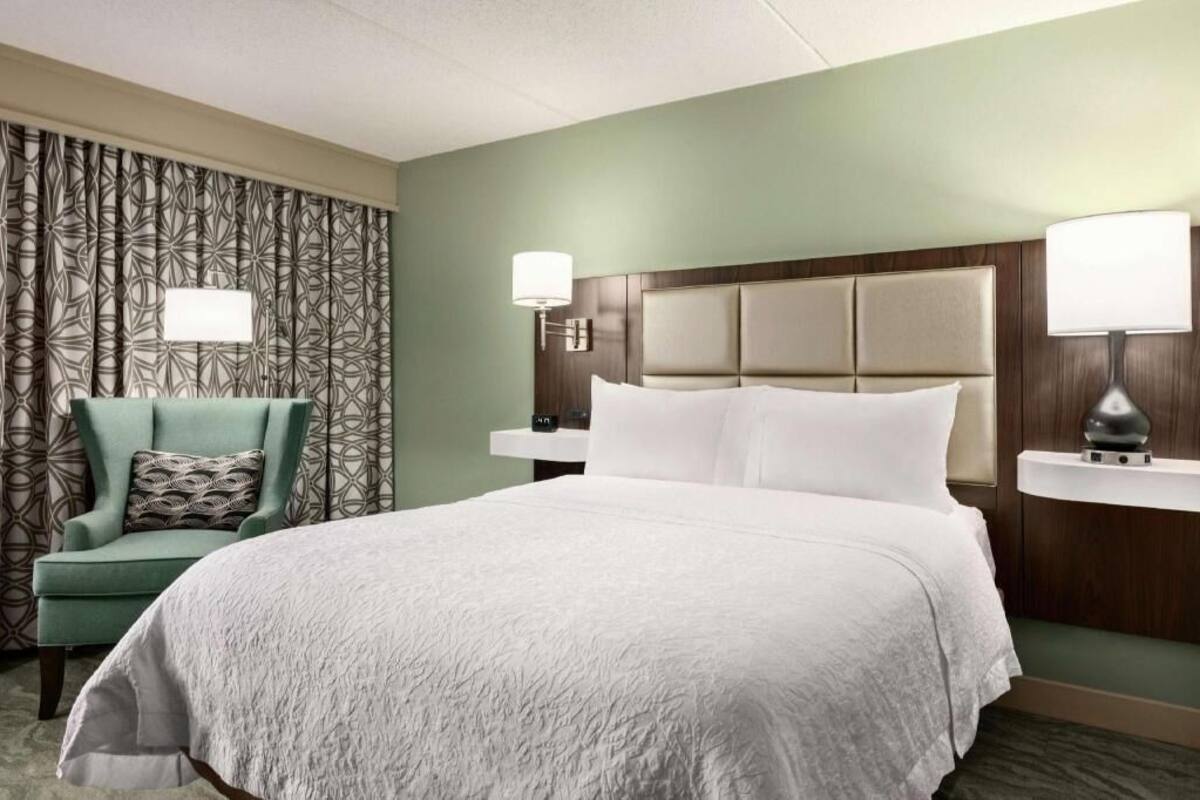 Quarto de hotel com uma cama grande, uma poltrona verde, uma mesa de cabeceira e cortina estampada.