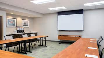 Sala de conferência com uma longa mesa de madeira, cadeiras, tela de projetor e um espaço para café.