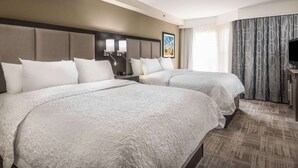 Quarto de hotel com duas camas, cabeceira, luminárias laterais, televisão e janela com cortinas.