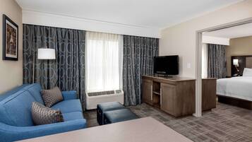 Quarto de hotel com sofá azul, televisão sobre um armário de madeira, cama e escrivaninha.