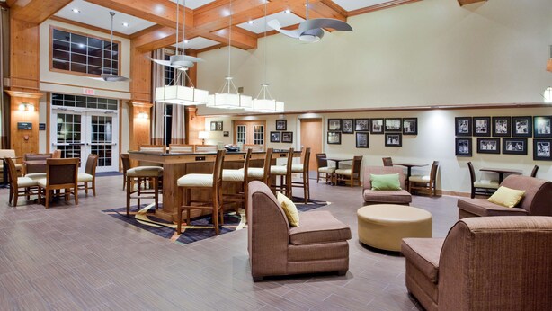 Um hall espaçoso com vigas de madeira no teto, área de bar central, diversas opções de assentos incluindo sofás e cadeiras, e quadros nas paredes.