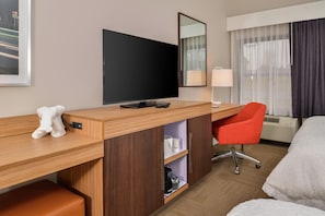 Quarto de hotel com escrivaninha de madeira, televisão, cadeira vermelha, cama e janela com cortinas.