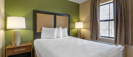 Quarto de hotel com uma cama grande, duas luminárias de cabeceira, uma mesinha de cabeceira com gaveta e uma janela com cortinas.