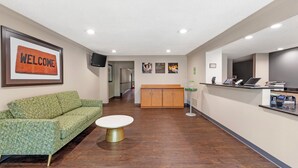 Uma área de recepção com balcão, um sofá, uma mesa de centro, TV na parede e um letreiro com a palavra 'WELCOME'.