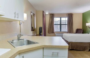 Quarto de hotel com cozinha compacta, cama, poltrona e janela com cortinas.