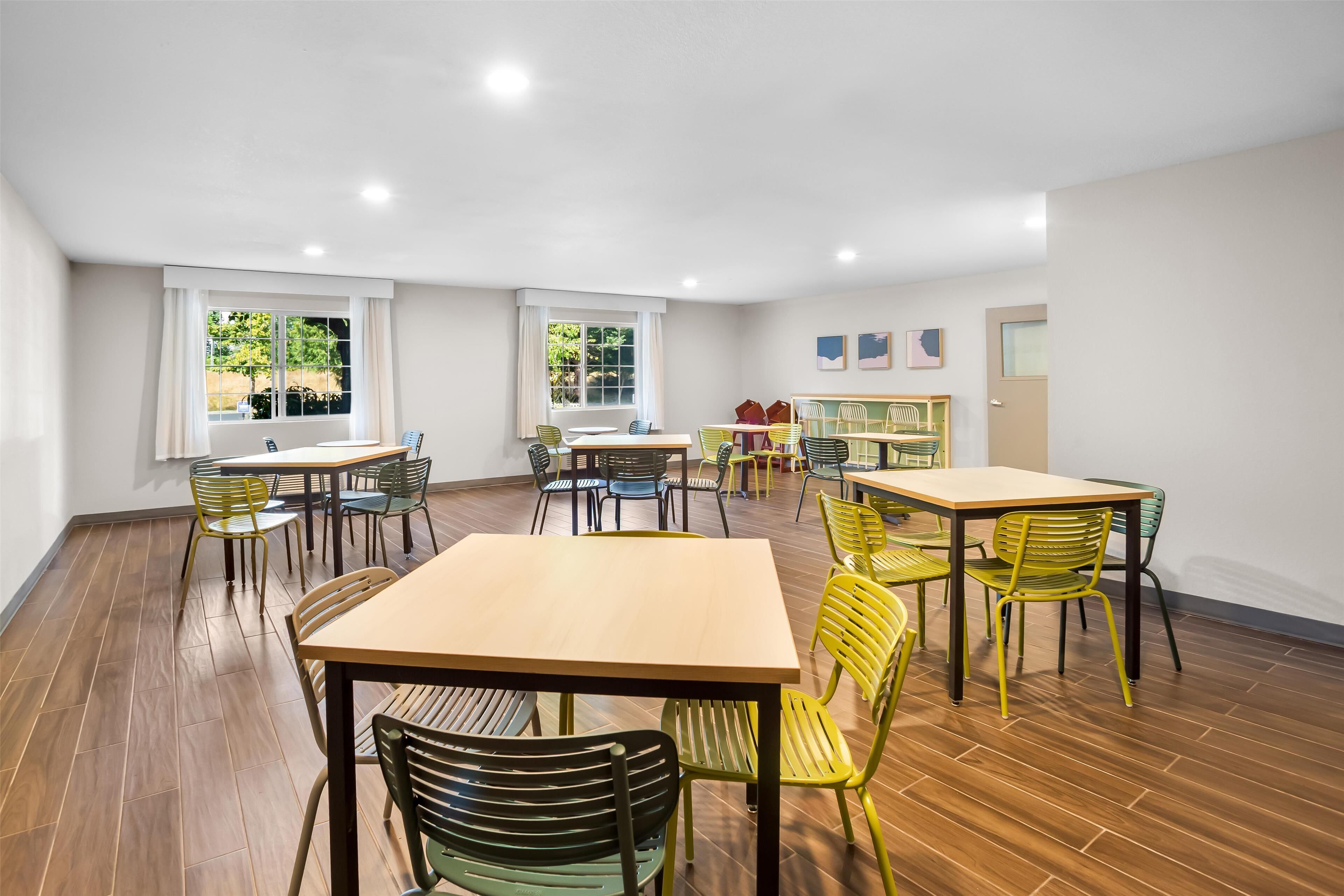 Um quarto espaçoso com várias mesas e cadeiras, janelas amplas com cortinas e uma porta.