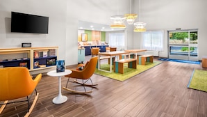 Um hall de hotel moderno com televisão de tela plana, área de recreação infantil, espaço de refeições com móveis de madeira e uma grande janela com vista para o exterior.