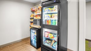 Uma cozinha compacta com duas geladeiras repletas de lanches e bebidas, um micro-ondas e uma pequena estante com temperos.