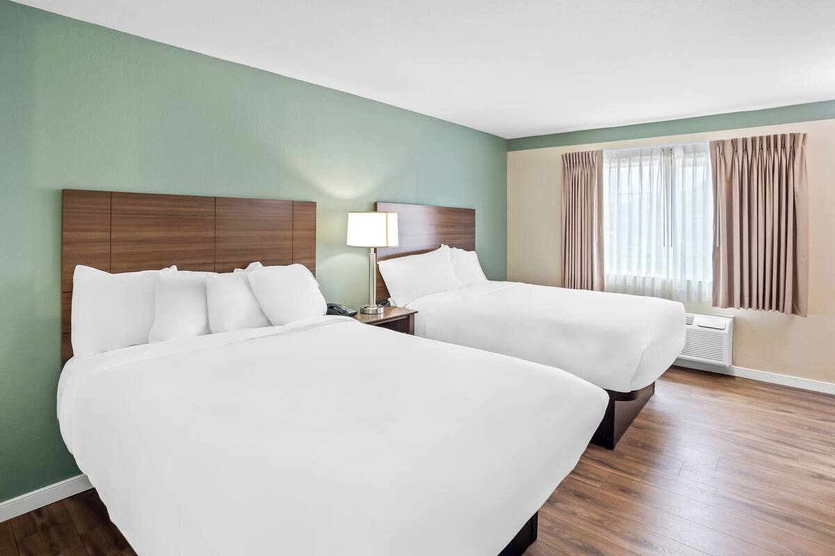 Quarto de hotel com duas camas, uma mesinha de cabeceira com luminária e uma janela com cortinas.