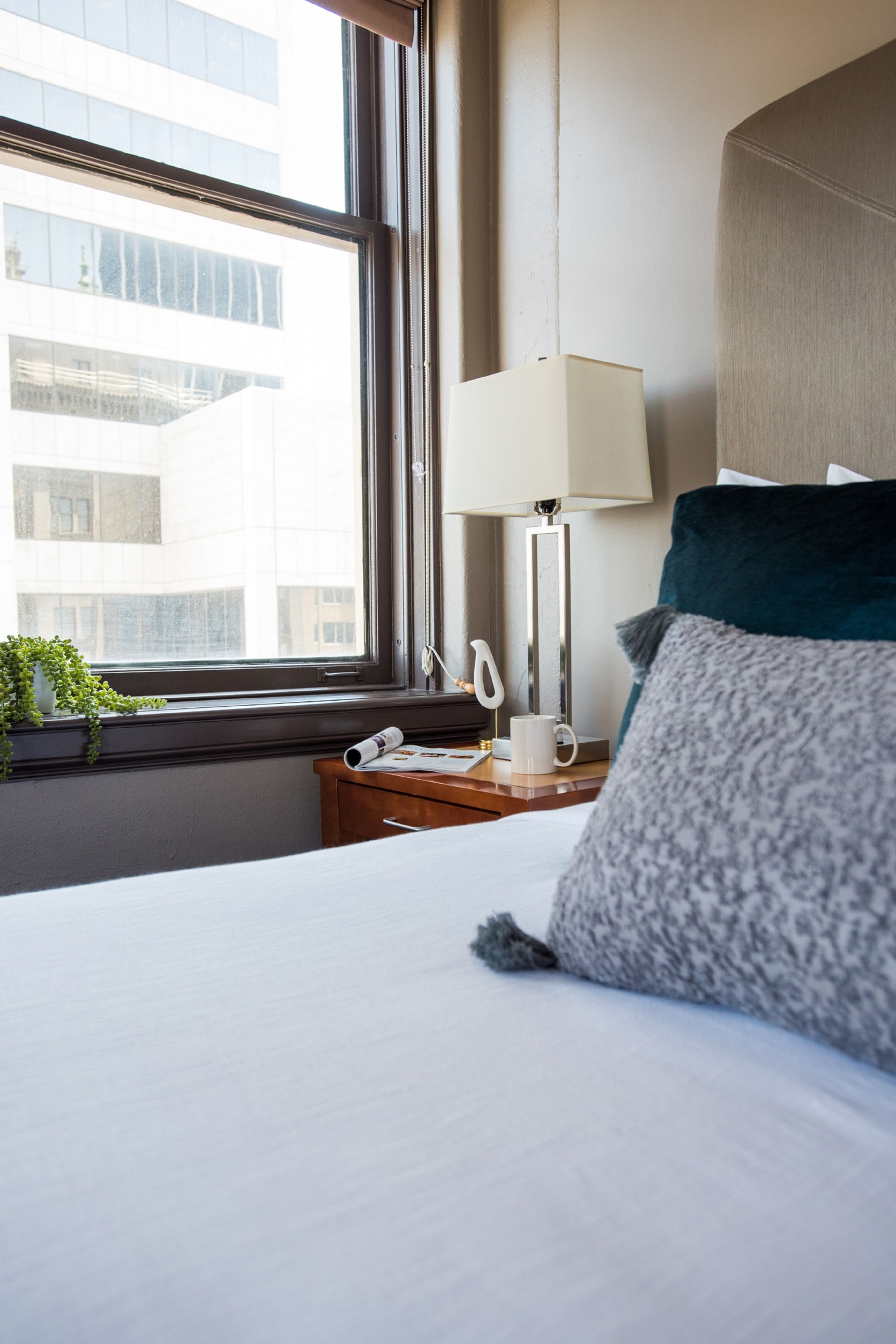 Quarto de hotel com cama, mesa de cabeceira com luminária, telefone e janela com vista para edifícios.