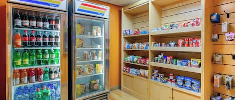 Uma loja de conveniência com dois refrigeradores de bebidas, prateleiras repletas de lanches e bebidas, e uma unidade de exposição de madeira com produtos embalados.