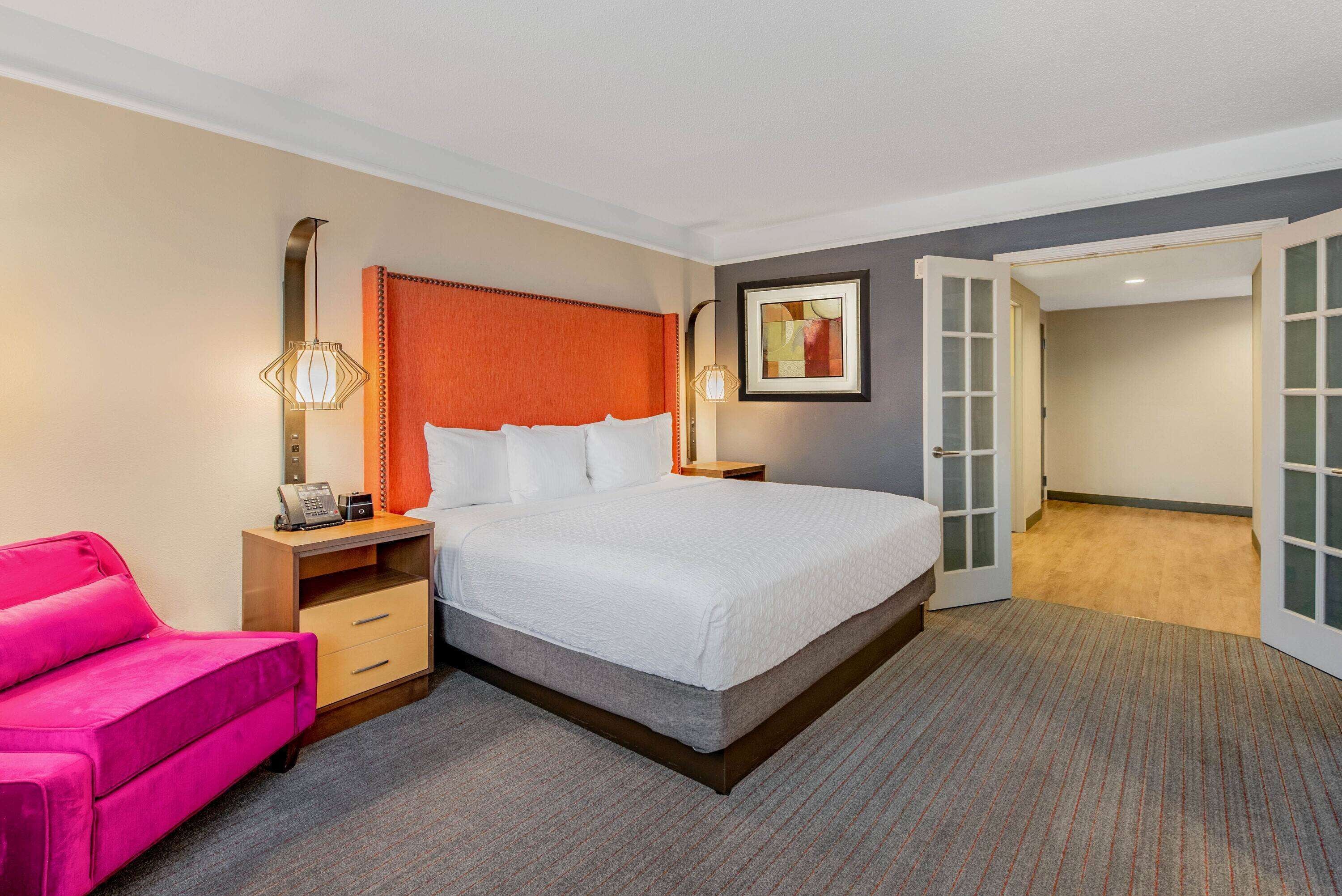 Quarto de hotel com uma cama, mesa de cabeceira, telefone, cadeira rosa e porta para outro cômodo.