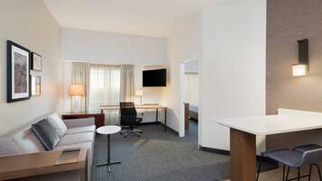 Quarto de hotel com sofá, escrivaninha, cadeira, televisão e luminária.
