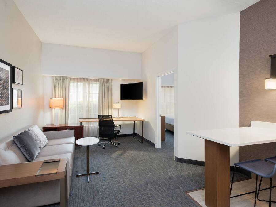 Quarto de hotel com sofá, escrivaninha, cadeira, televisão e luminária.