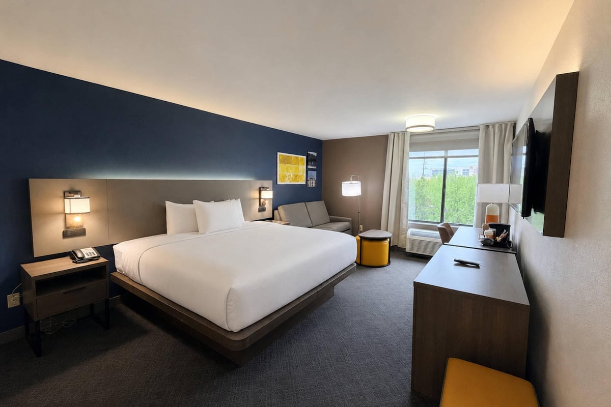 Quarto de hotel com cama grande, mesas de cabeceira, escrivaninha, cadeira e janela com cortinas.