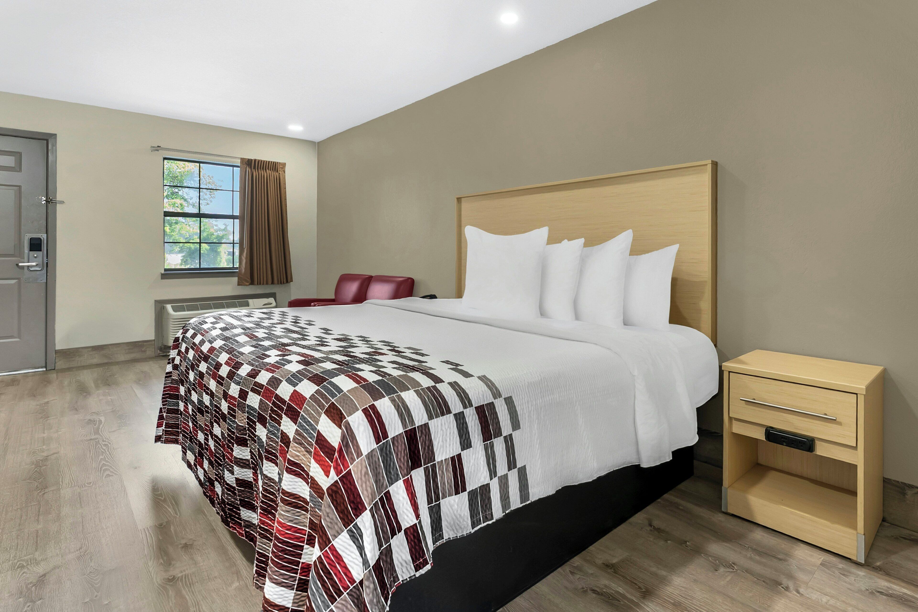 Quarto de hotel com uma cama grande, mesa de cabeceira com gavetas, janela com cortinas e um espaço de estar com cadeiras.