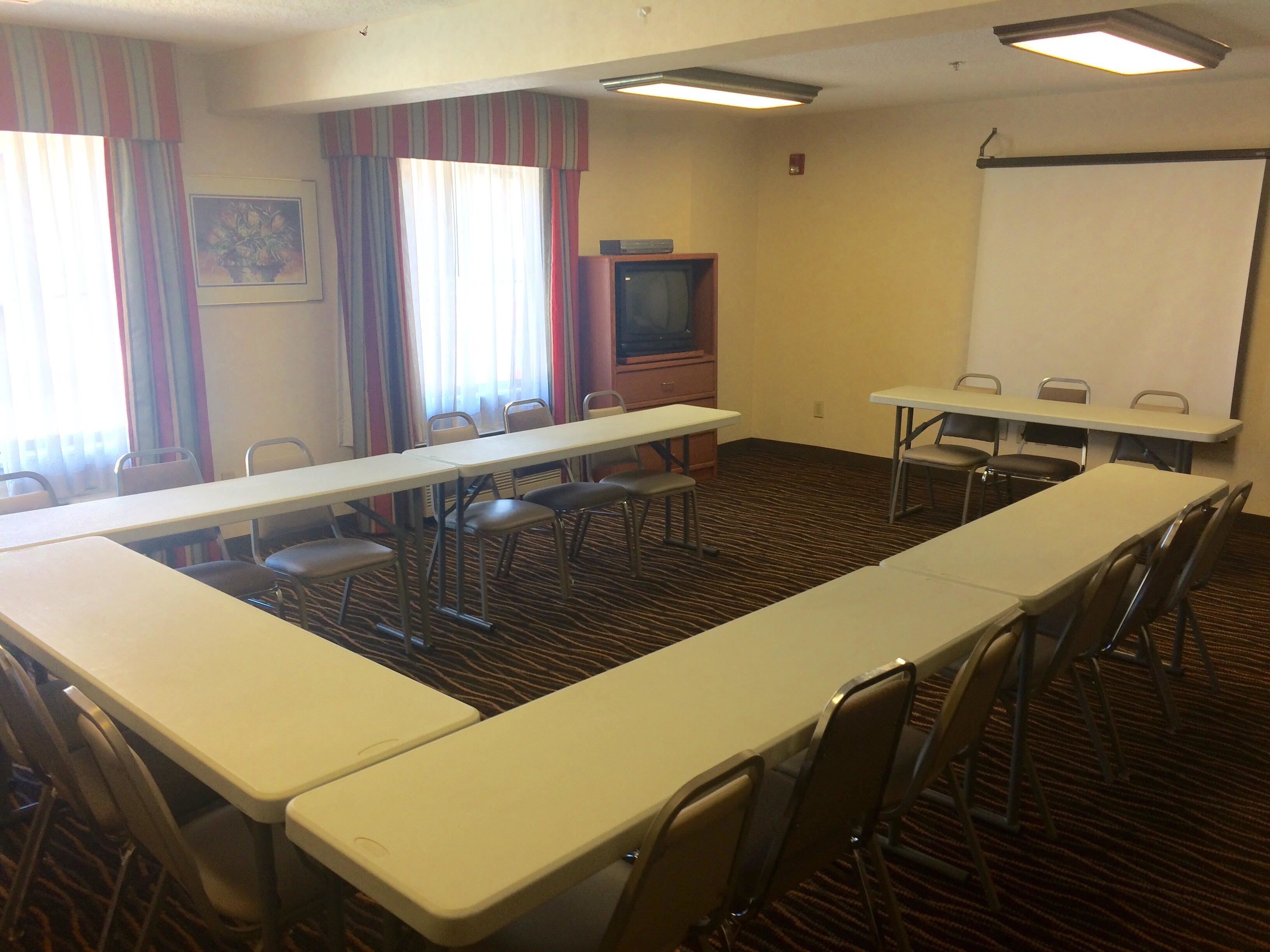 Uma sala de conferências com mesas compridas e cadeiras, uma tela de projetor e uma televisão.